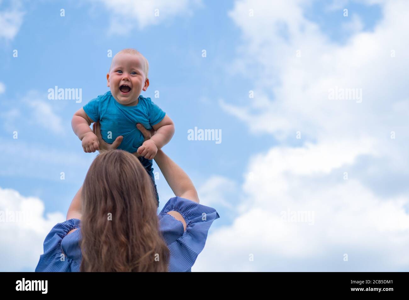 Mom lifting baby high into blue cloudy sky. Boy smiling and looking at ...