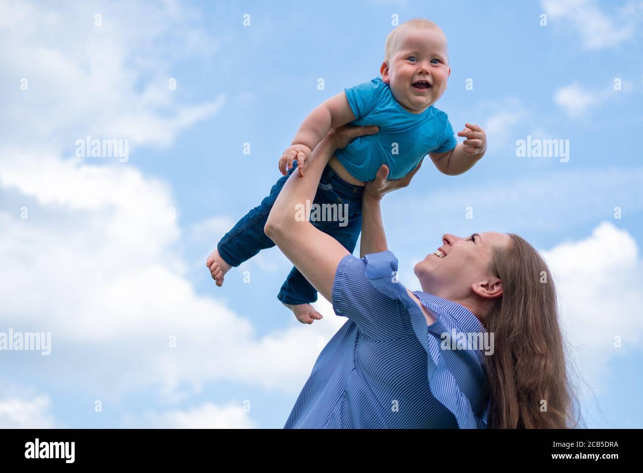 Mom lifting baby high into blue cloudy sky. Boy smiling and looking at ...