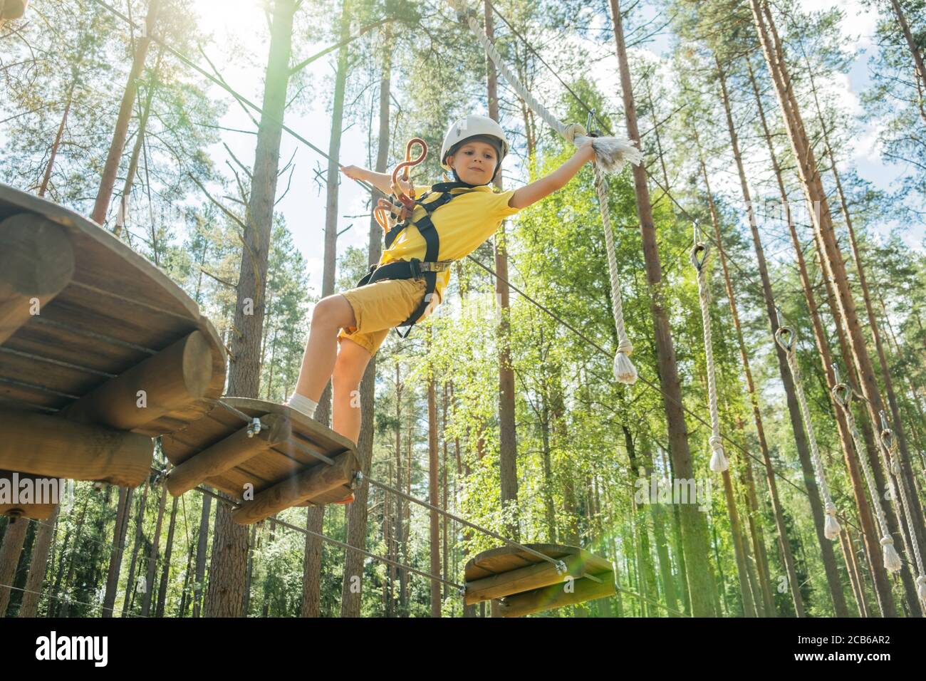 Little boy with climbing gear climbing rope trail between pine trees in ...