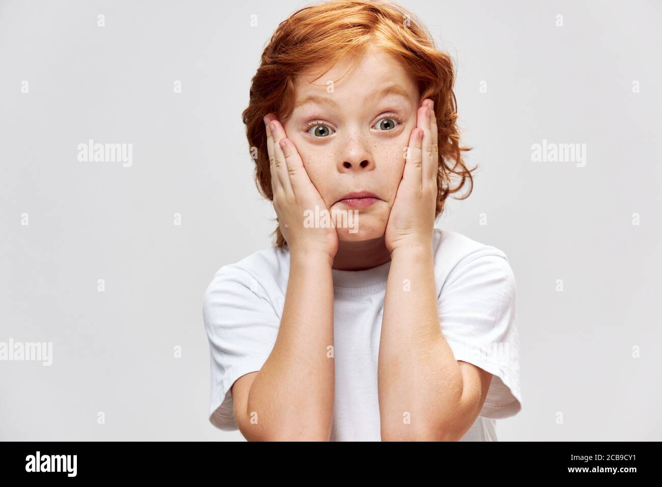 Red-haired boy holding his face in a white T-shirt surprised look close ...