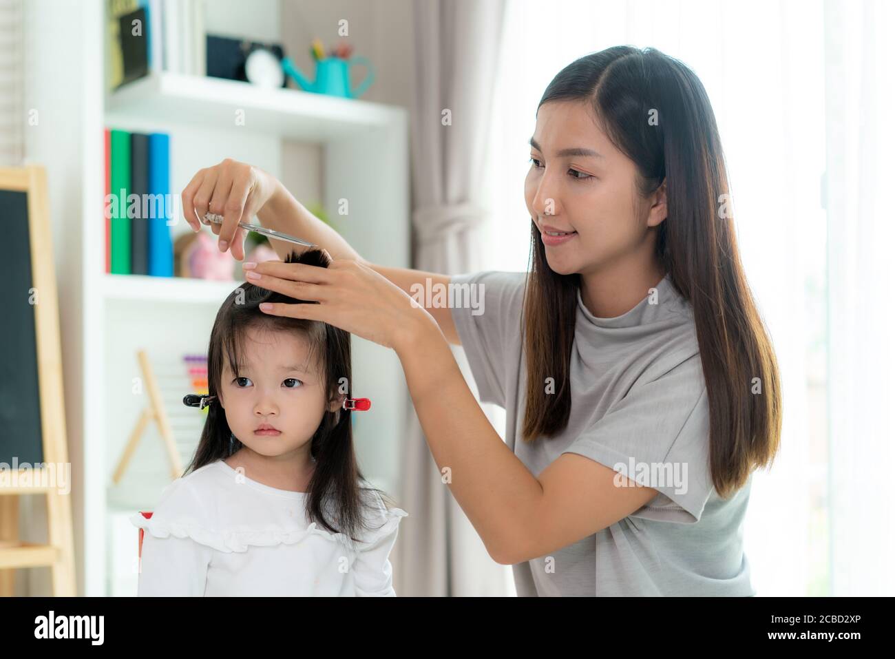 Asian Mother Cutting Hair To Her Daughter In Living Room At Home While