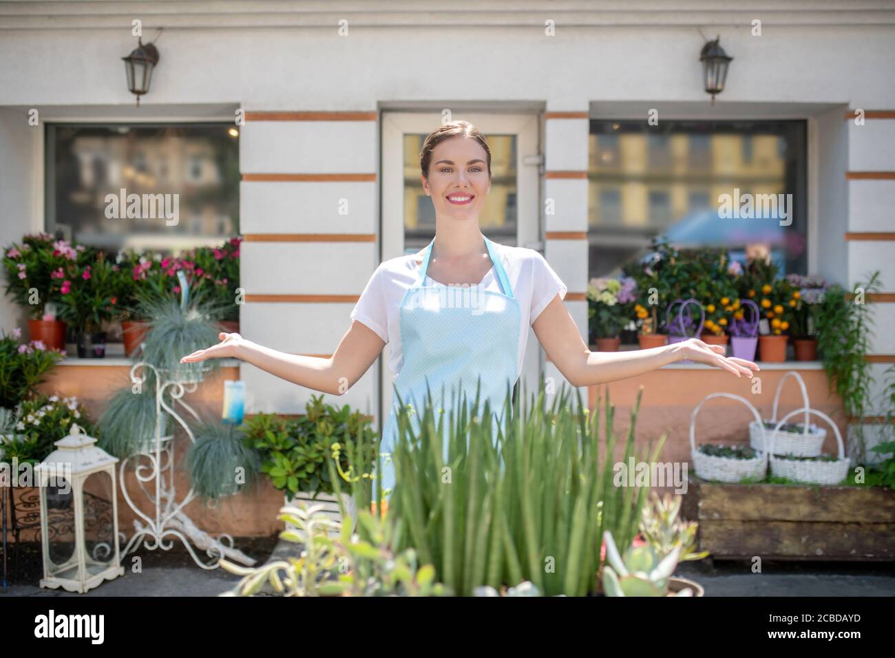 Pretty smiling female in apron standing in front of table with flowers ...