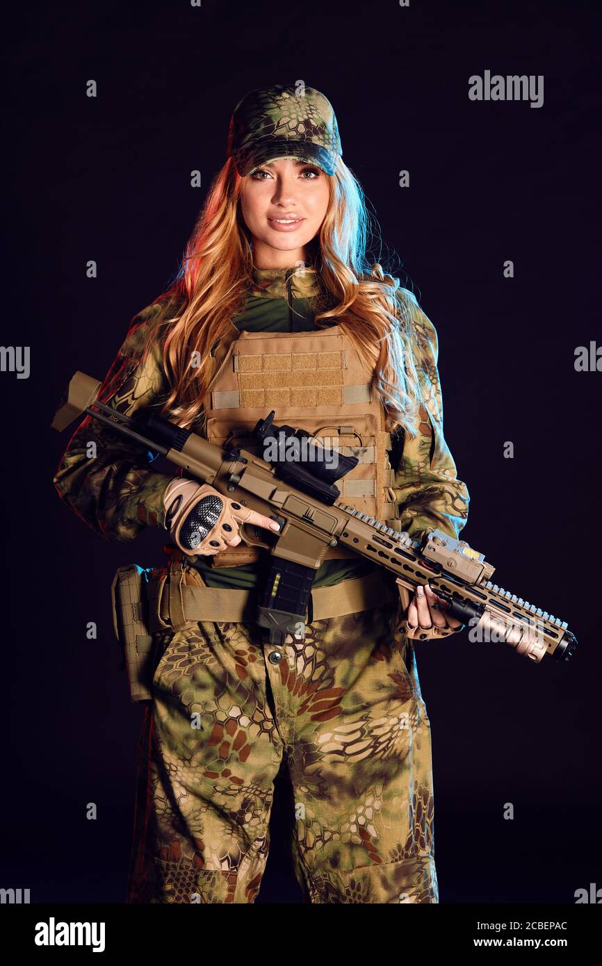 Sniper woman in ghillie suit, cap and plate carrier holds airsoft rifle over black background ...