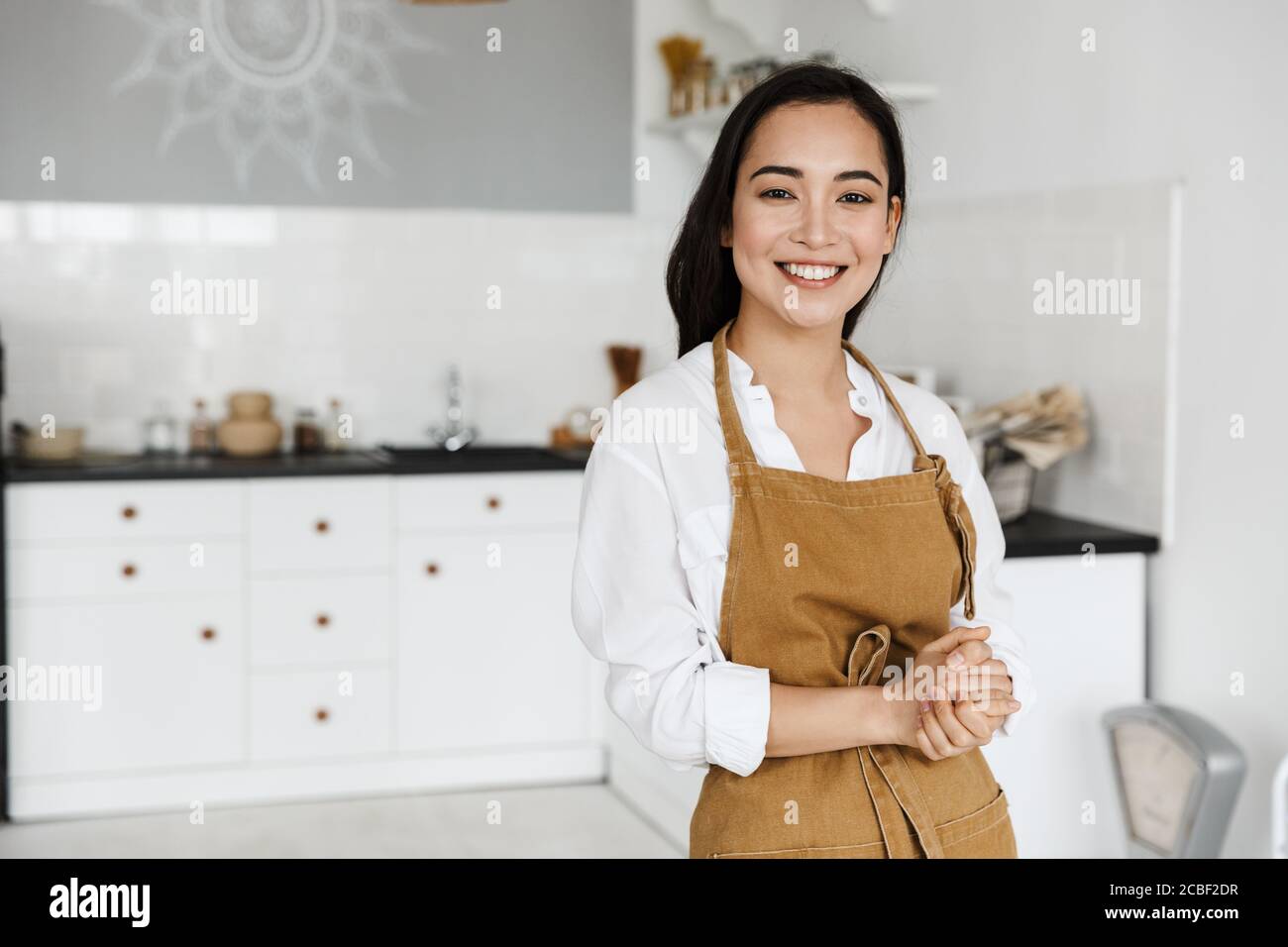 Close up of a confident smiling young asian woman wearing apron standing in the kitchen at home ...