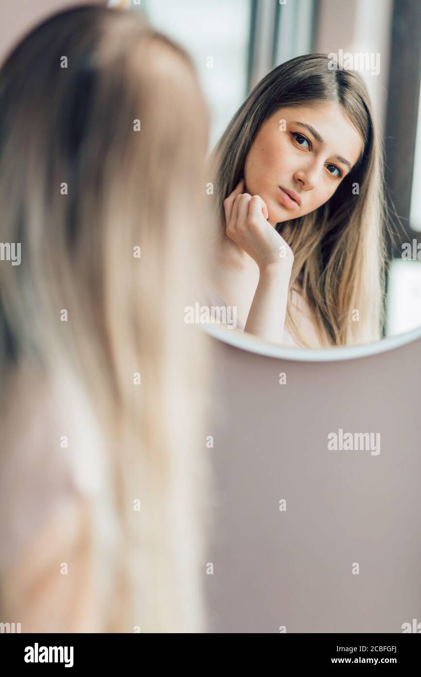 awesome girl with long fair hair looking in mirror at home. close up ...