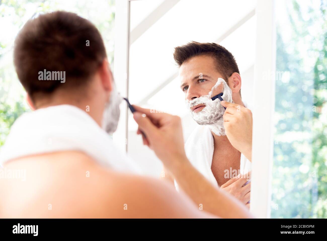 Shot of handsome man with towel around his neck using shaving foam while shawing in front of ...