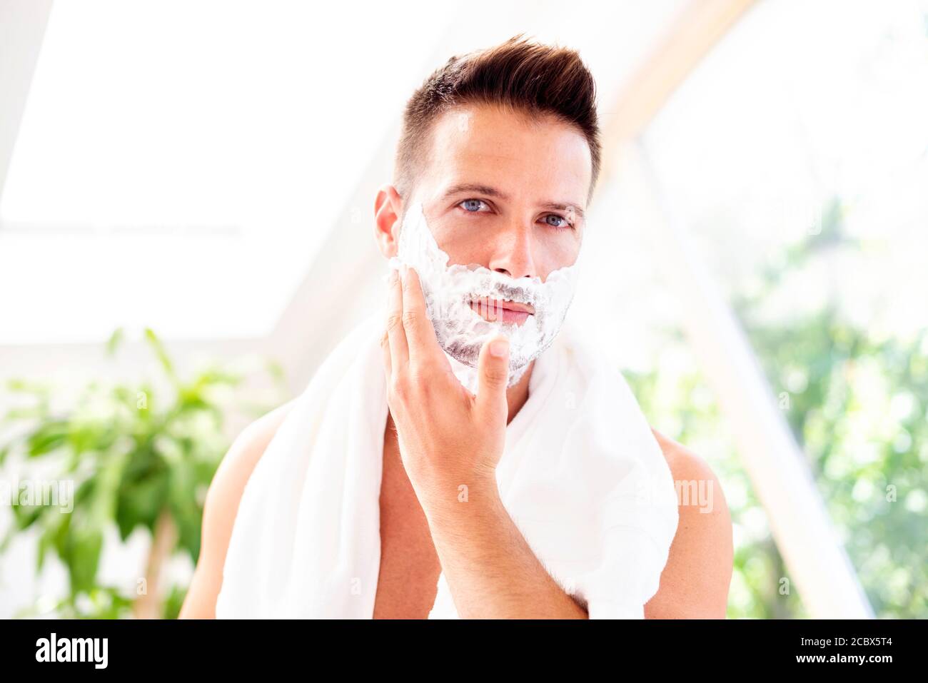 Shot of handsome man with towel around his neck using shaving foam before shaving while standing ...