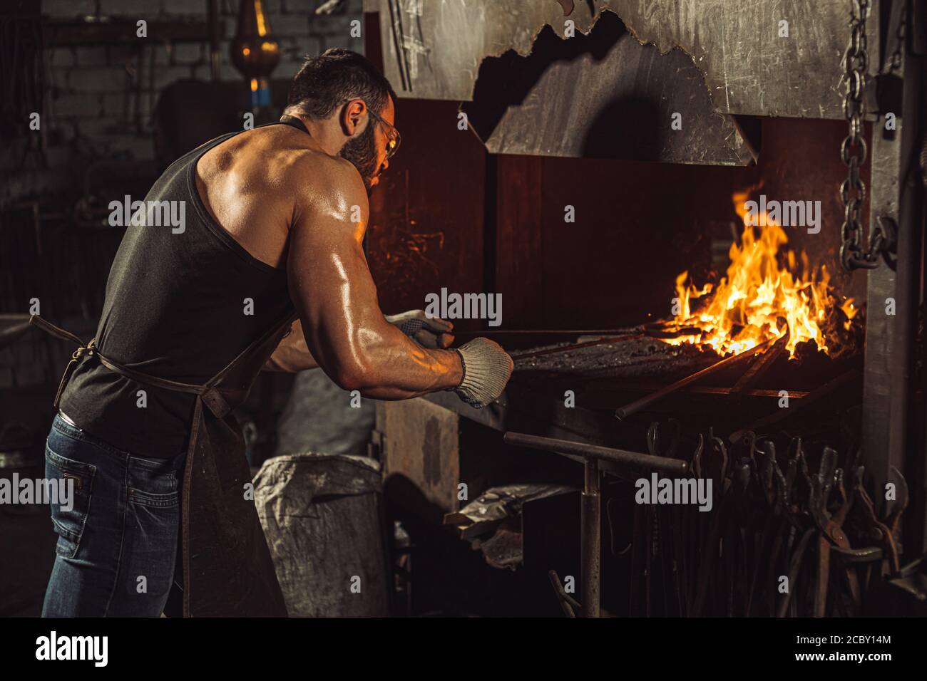 young caucasian strong man in black apron heating steel piece in ...