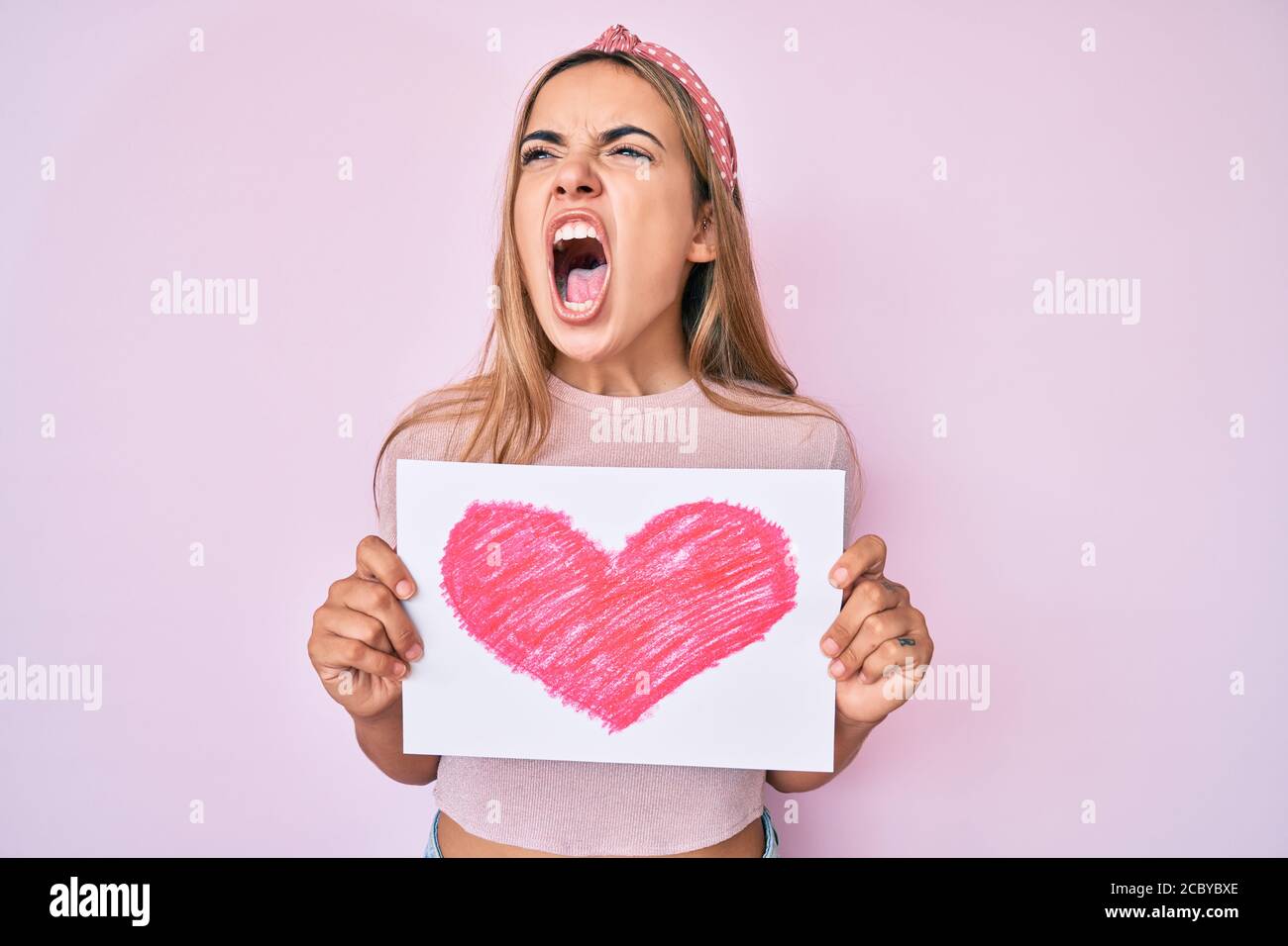 Young beautiful blonde woman holding heart draw angry and mad screaming ...