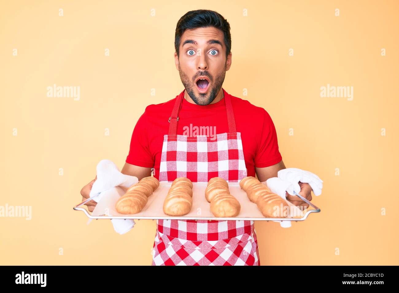 Young hispanic man wearing baker apron holding homemade bread afraid ...