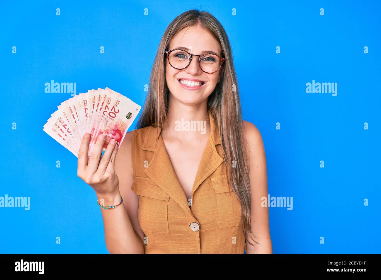 Young blonde girl holding israeli shekels looking positive and happy ...