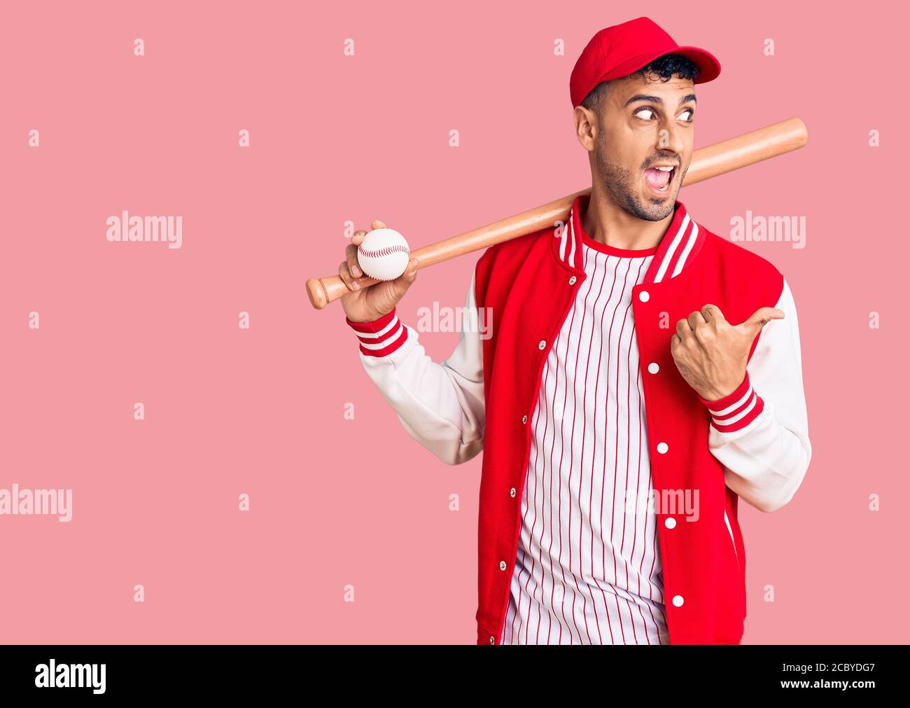 Young hispanic man playing baseball holding bat and ball pointing thumb ...