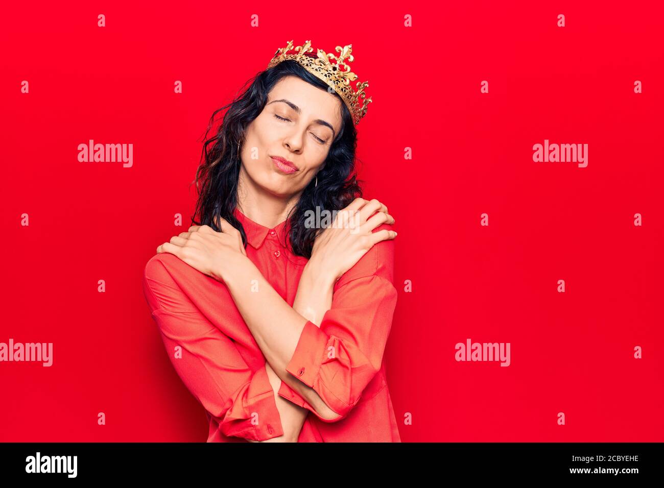 Young beautiful hispanic woman wearing princess crown hugging oneself ...