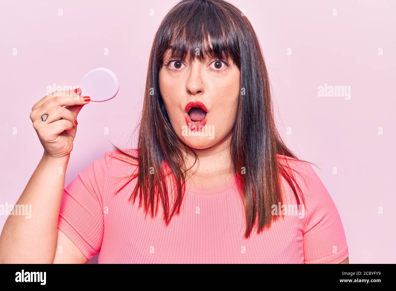 Young plus size woman holding makeup sponge scared and amazed with open ...