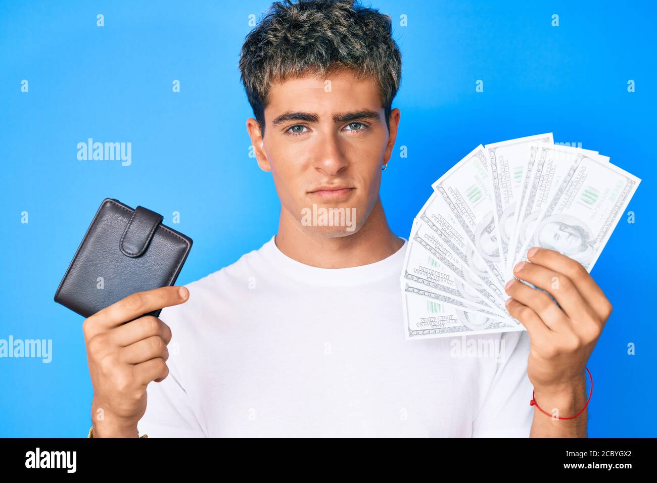 Young handsome man holding wallet and dollars banknotes skeptic and ...