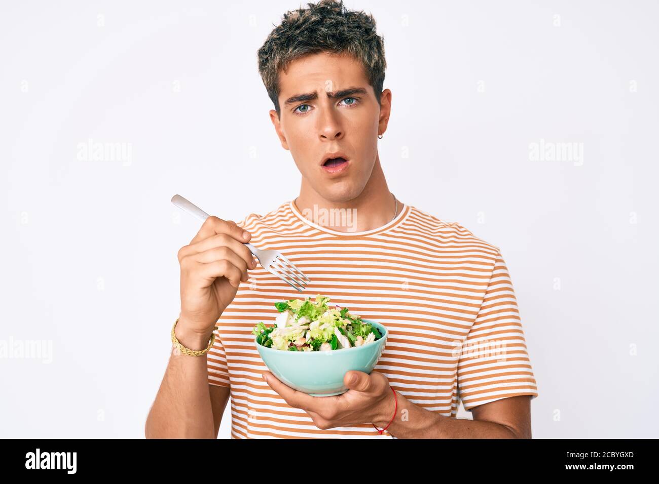 Young handsome man eating salad in shock face, looking skeptical and ...