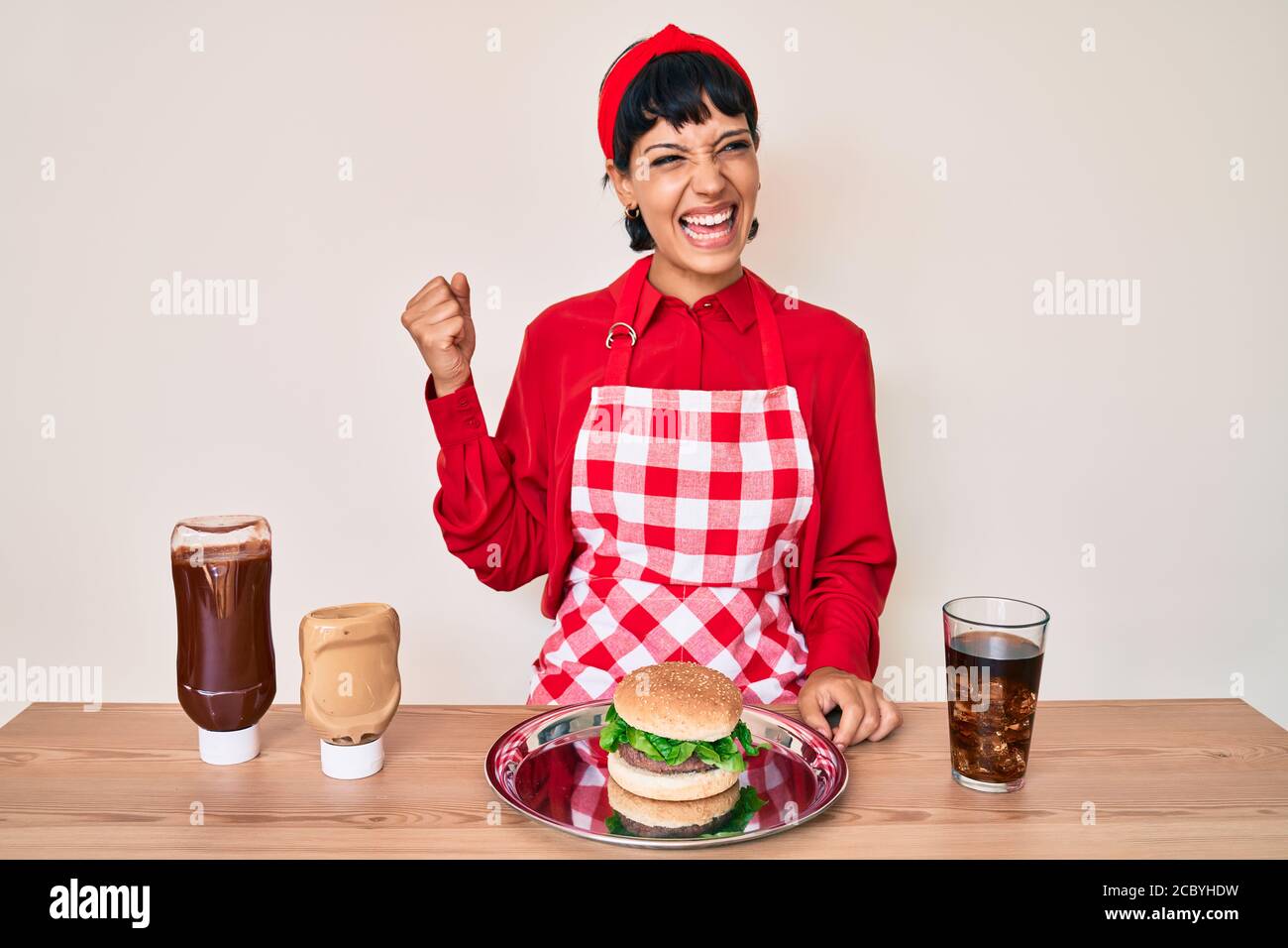 Beautiful brunettte woman cooking tasty hamburguer screaming proud, celebrating victory and ...