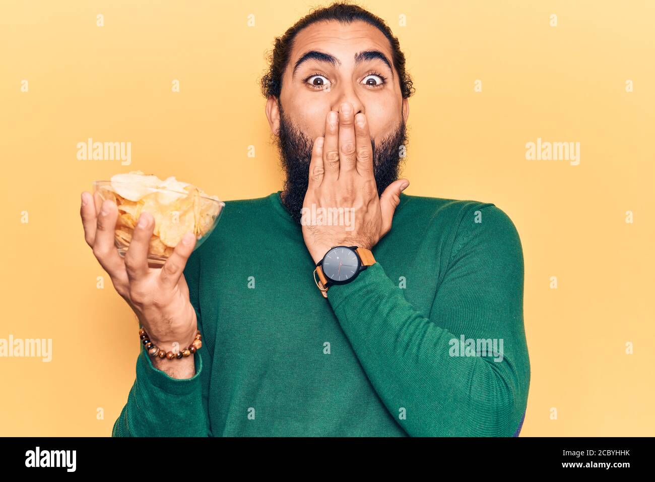 Young arab man holding potato chip covering mouth with hand, shocked ...