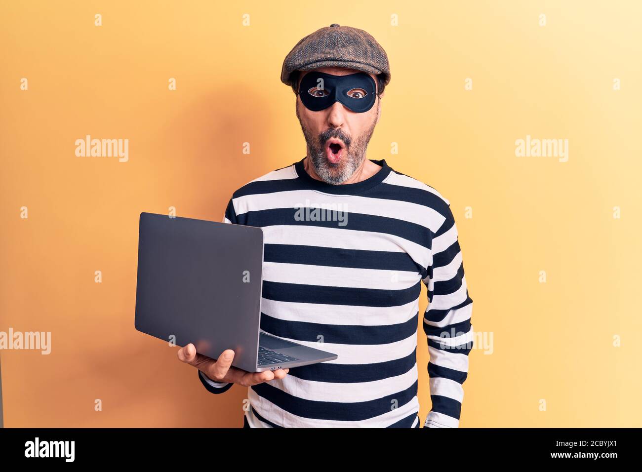 Middle age handsome burglar man wearing cap and mask using laptop over ...