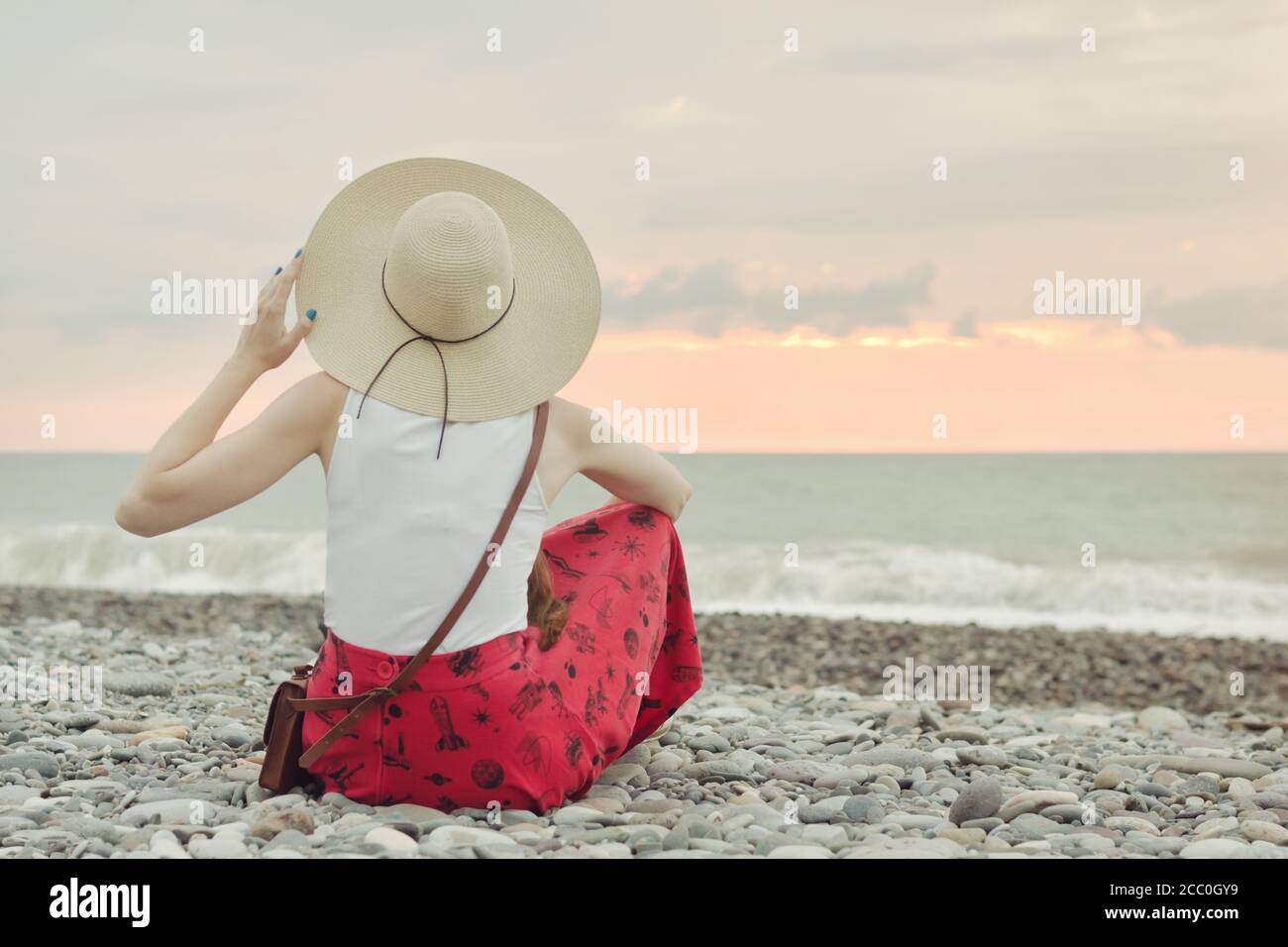 Girl in a hat sits on a pebble beach. Back view. Sunset time Stock ...