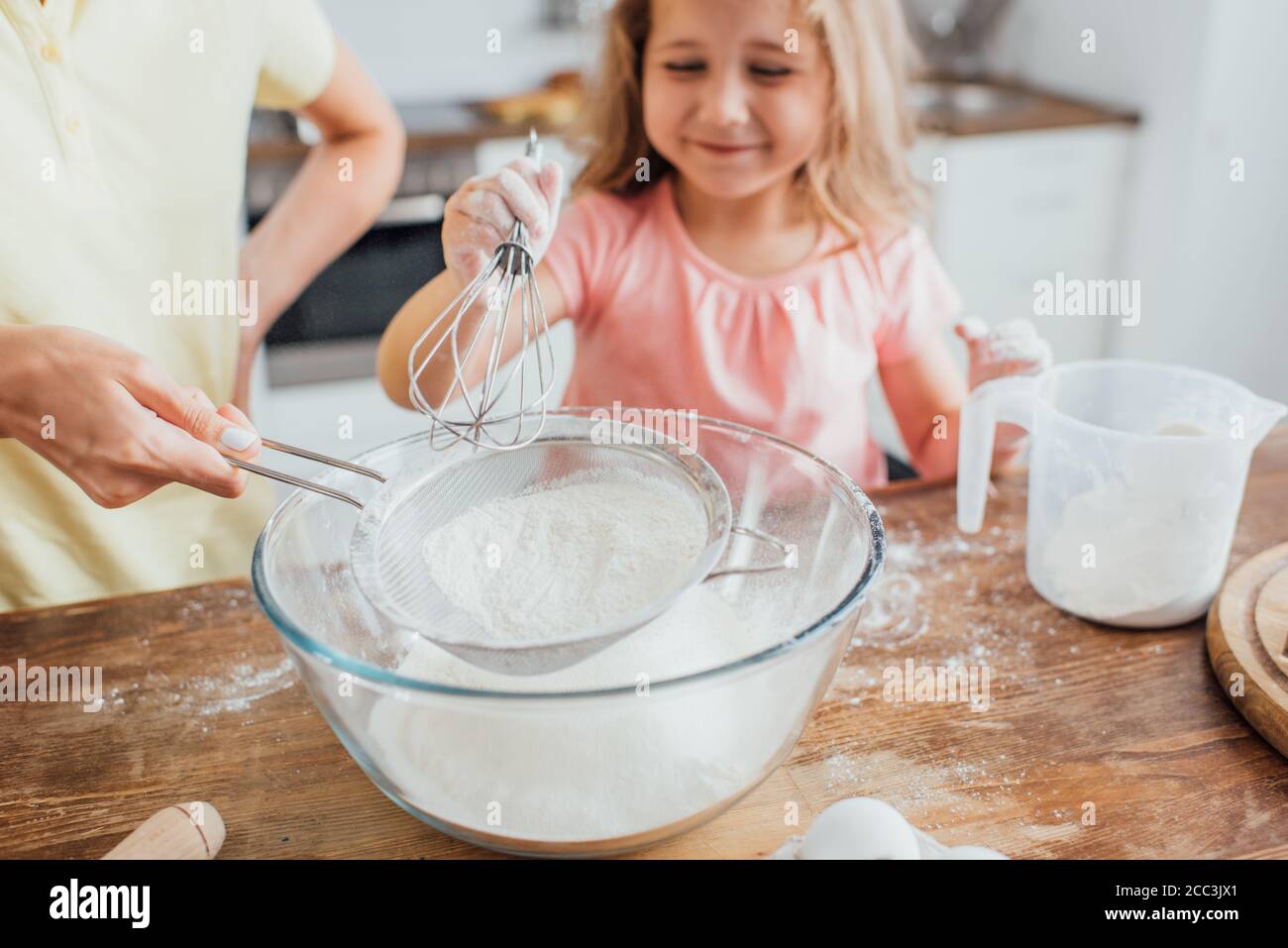 cropped view of mother with sieve and daughter with whisk sieving flour ...
