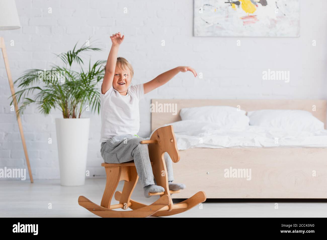 excited kid riding rocking horse with raised hands near potted plant ...