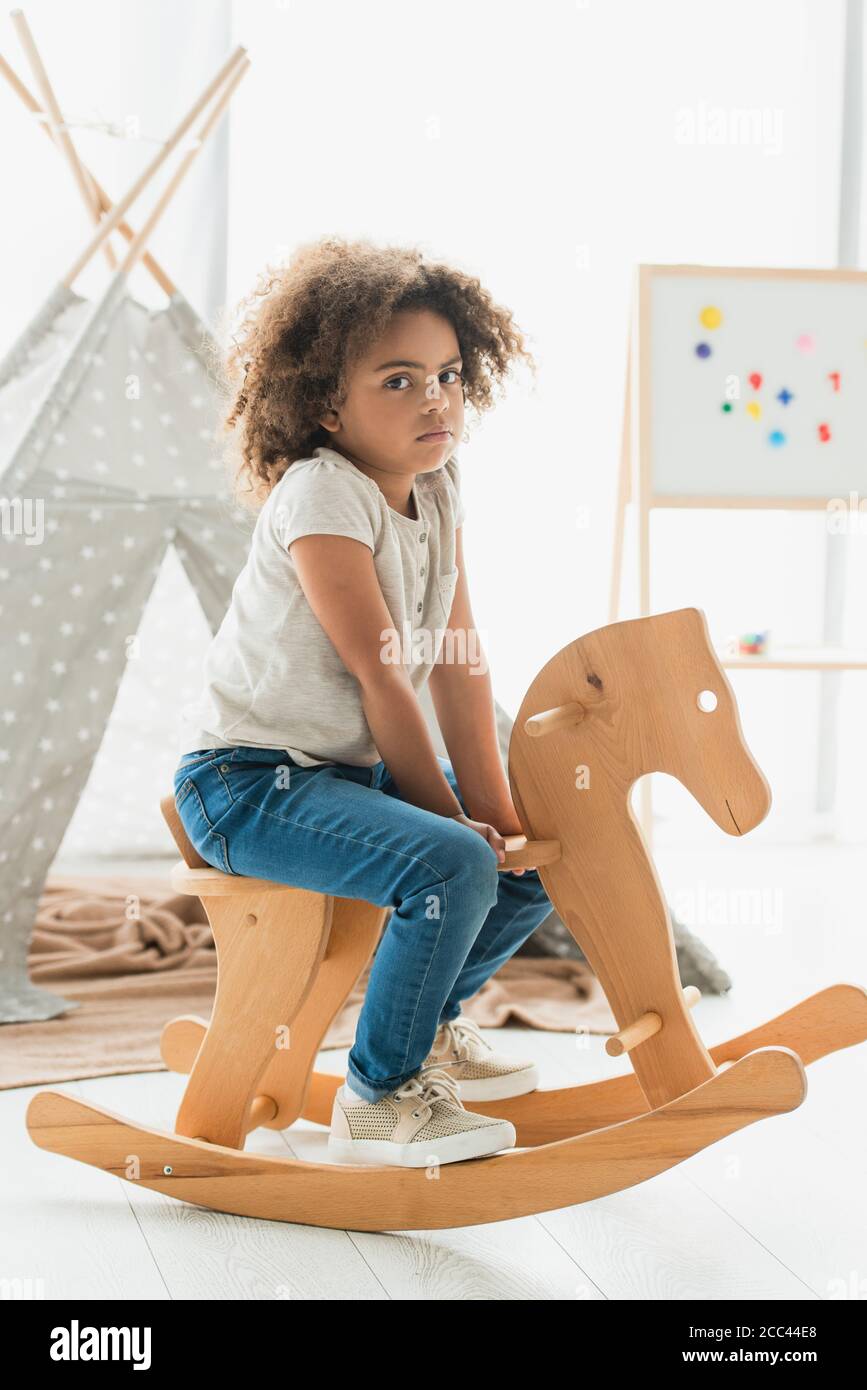 curly african american kid sitting on wooden rocking horse at home ...