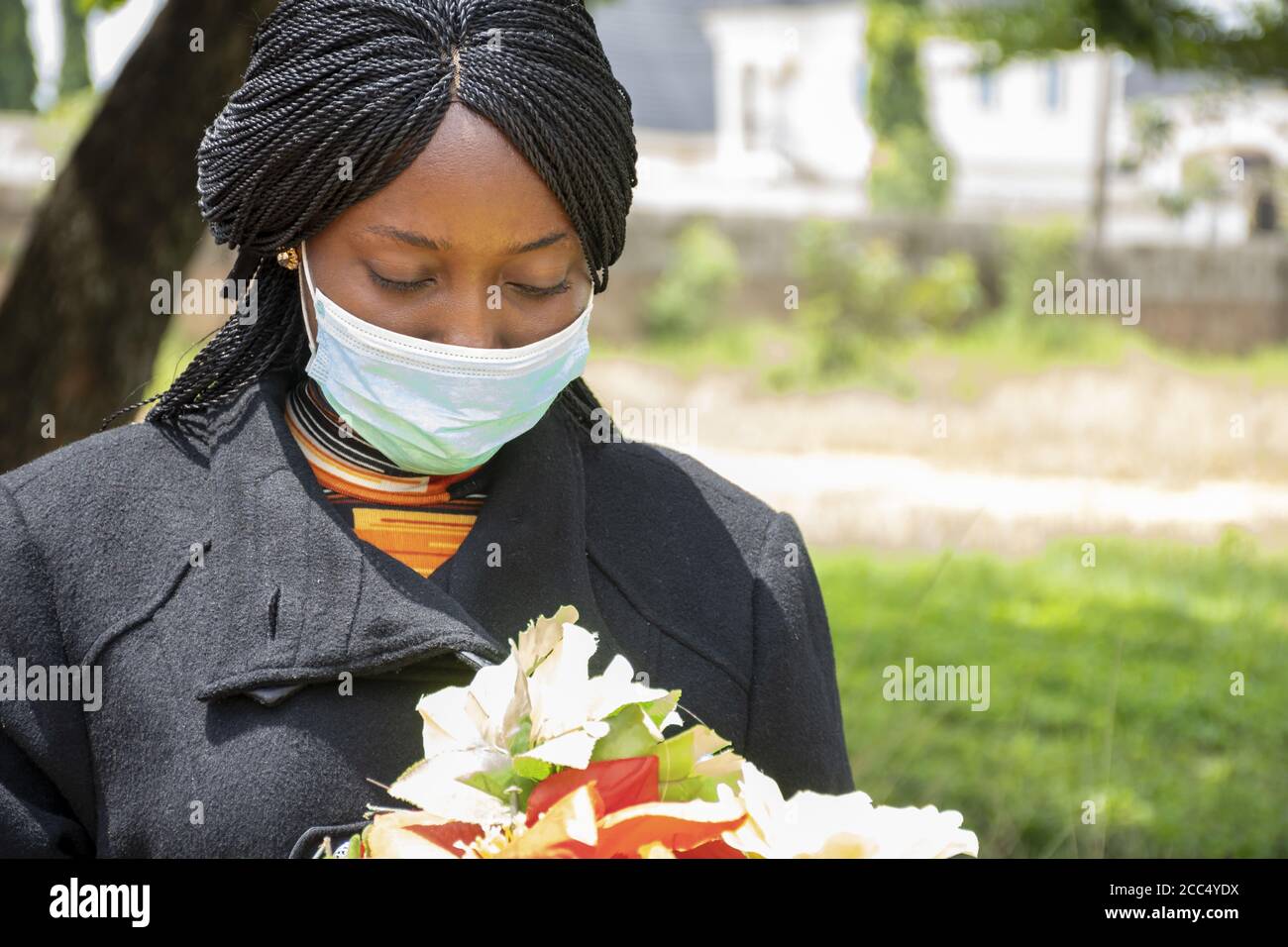 Shot of a black female mourning, wearing a mask and holding flowers ...