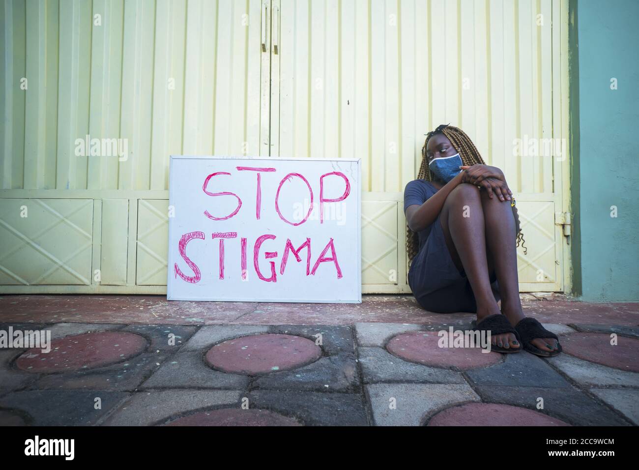 Young black female with a medical mask sitting next to a sign with text ...