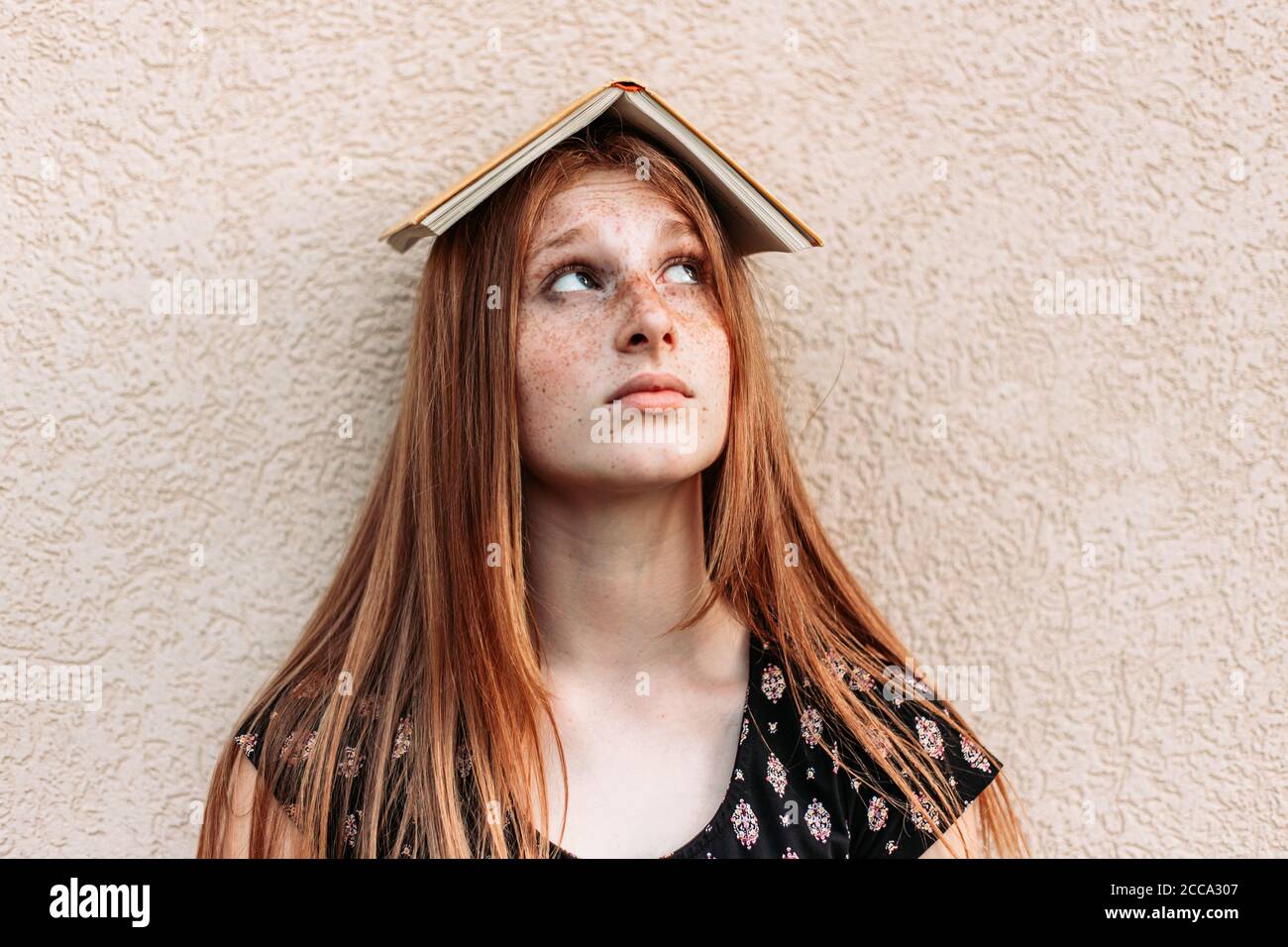 Teen ginger student girl with a book on top of her head, looking at it ...