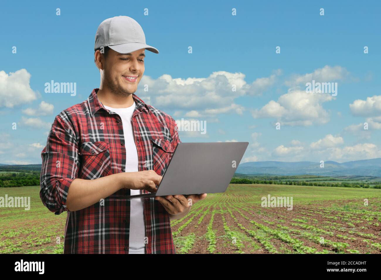 Young male farmer in a shirt holding a laptop computer on a field with ...