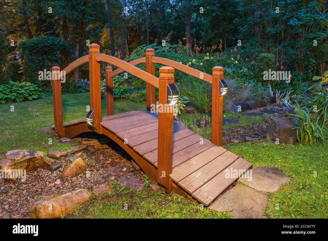 Footbridge over stream and pond at dusk bordered by perennial plants ...