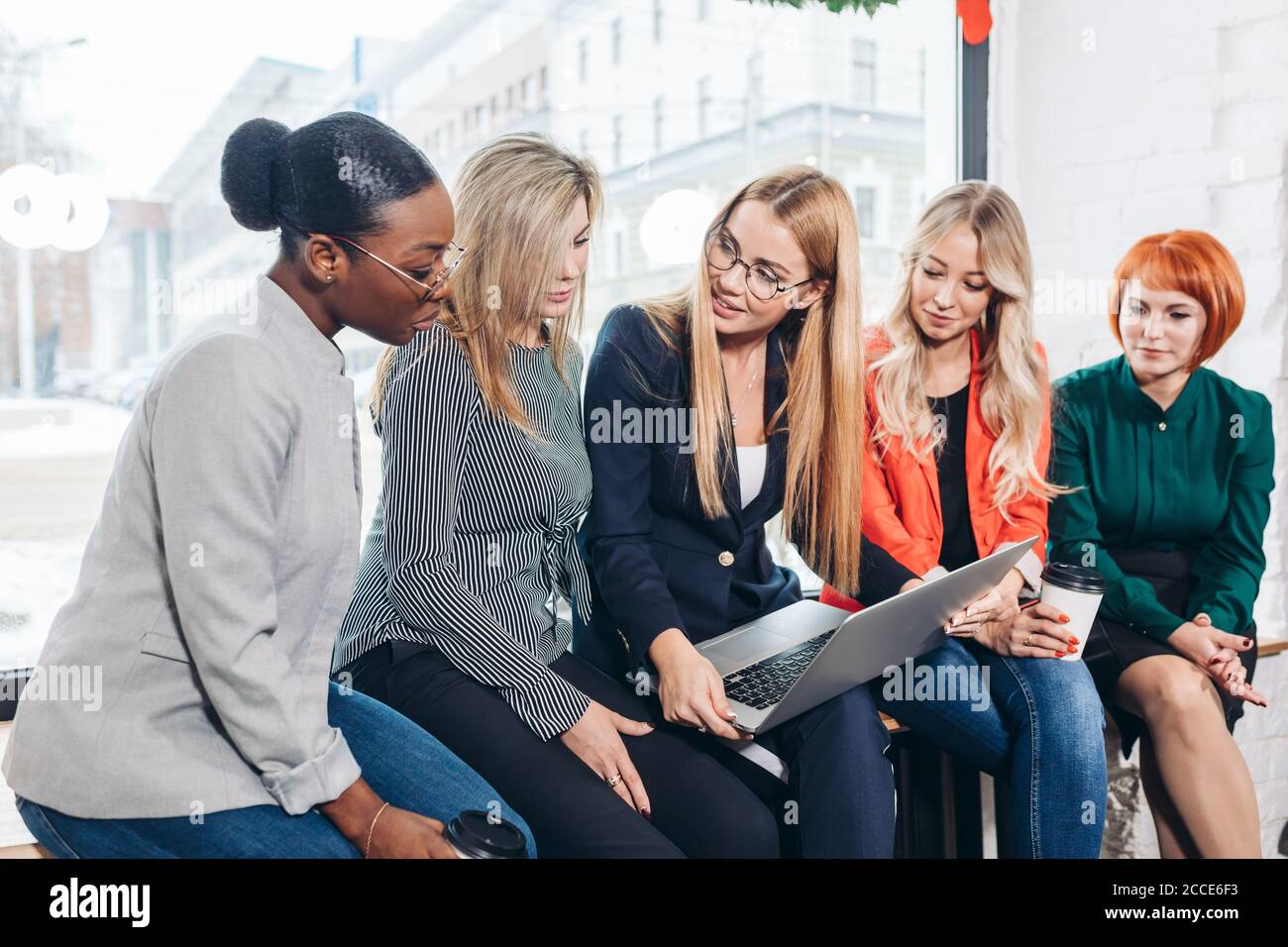 Multiracial group of female students sitting together at a window sill with coffee and laptop ...