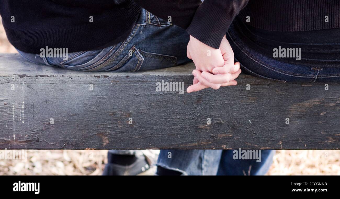A couple in jeans, sitting on a park bench, secretly hold hands behind ...