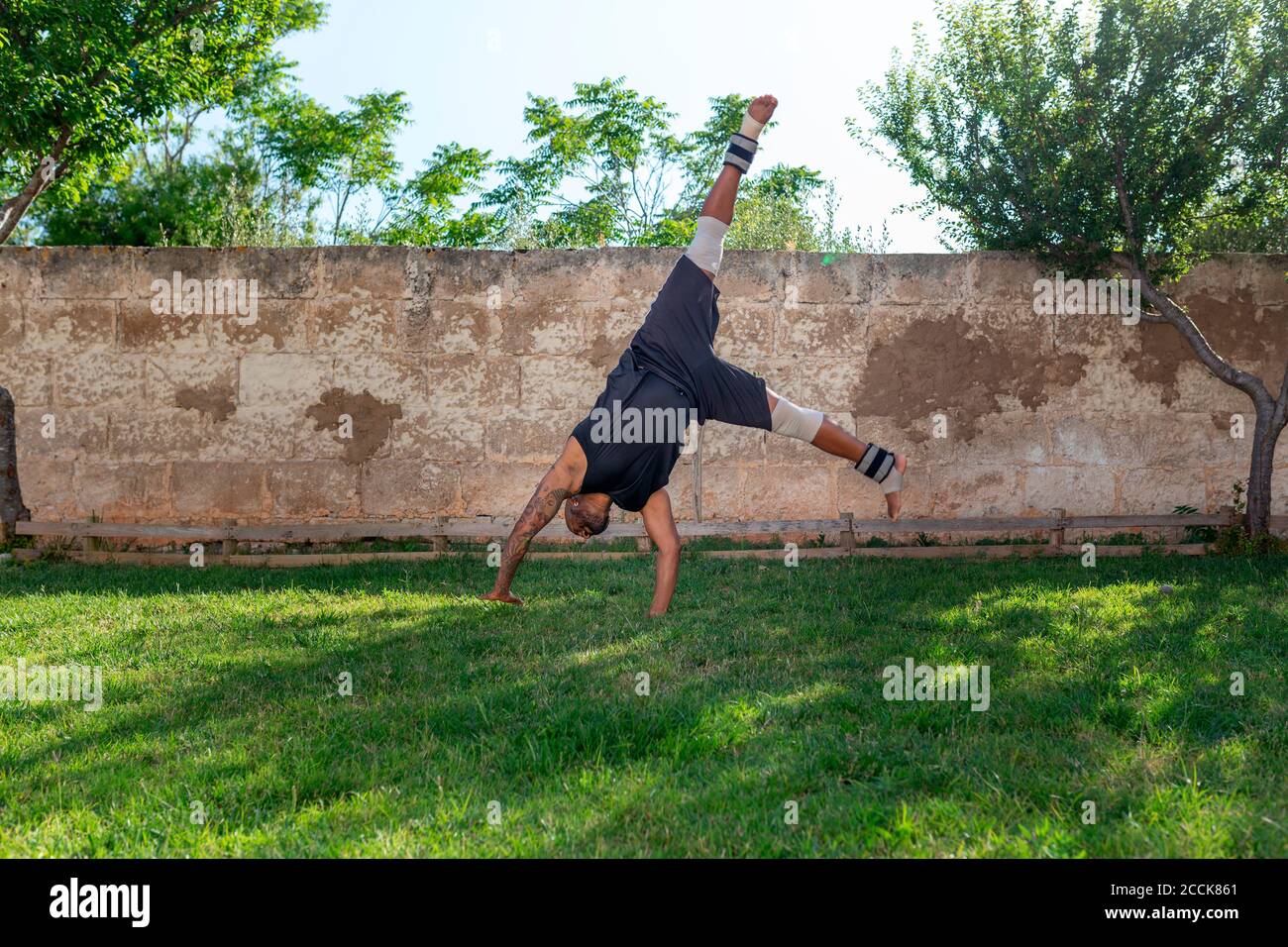 Mature man practicing cart wheel on grassy land against wall in yard ...