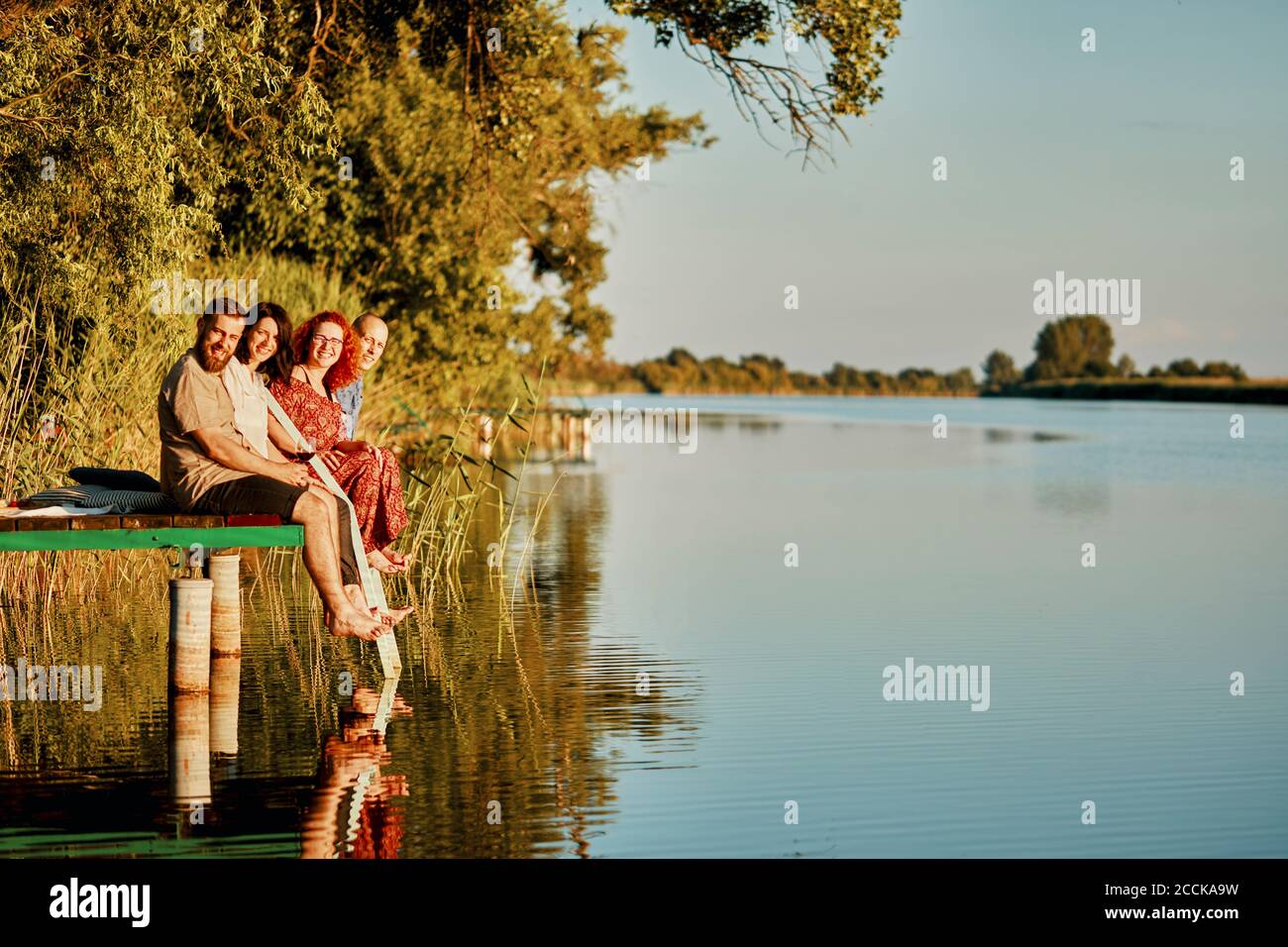 Portrait of smiling friends reflected in water sitting on jetty at a ...