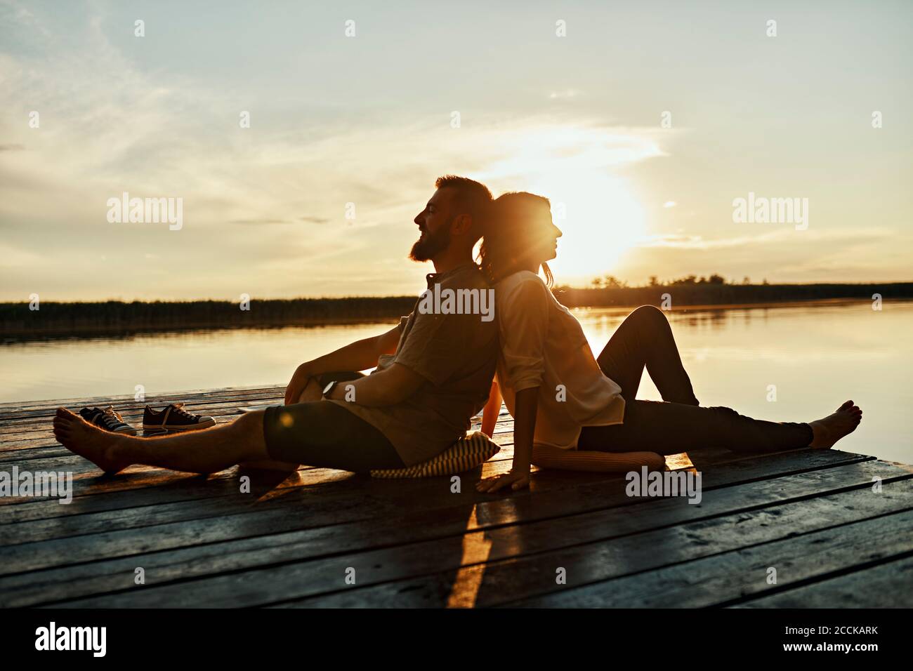 Couple sitting back to back on jetty at a lake at sunset Stock Photo ...