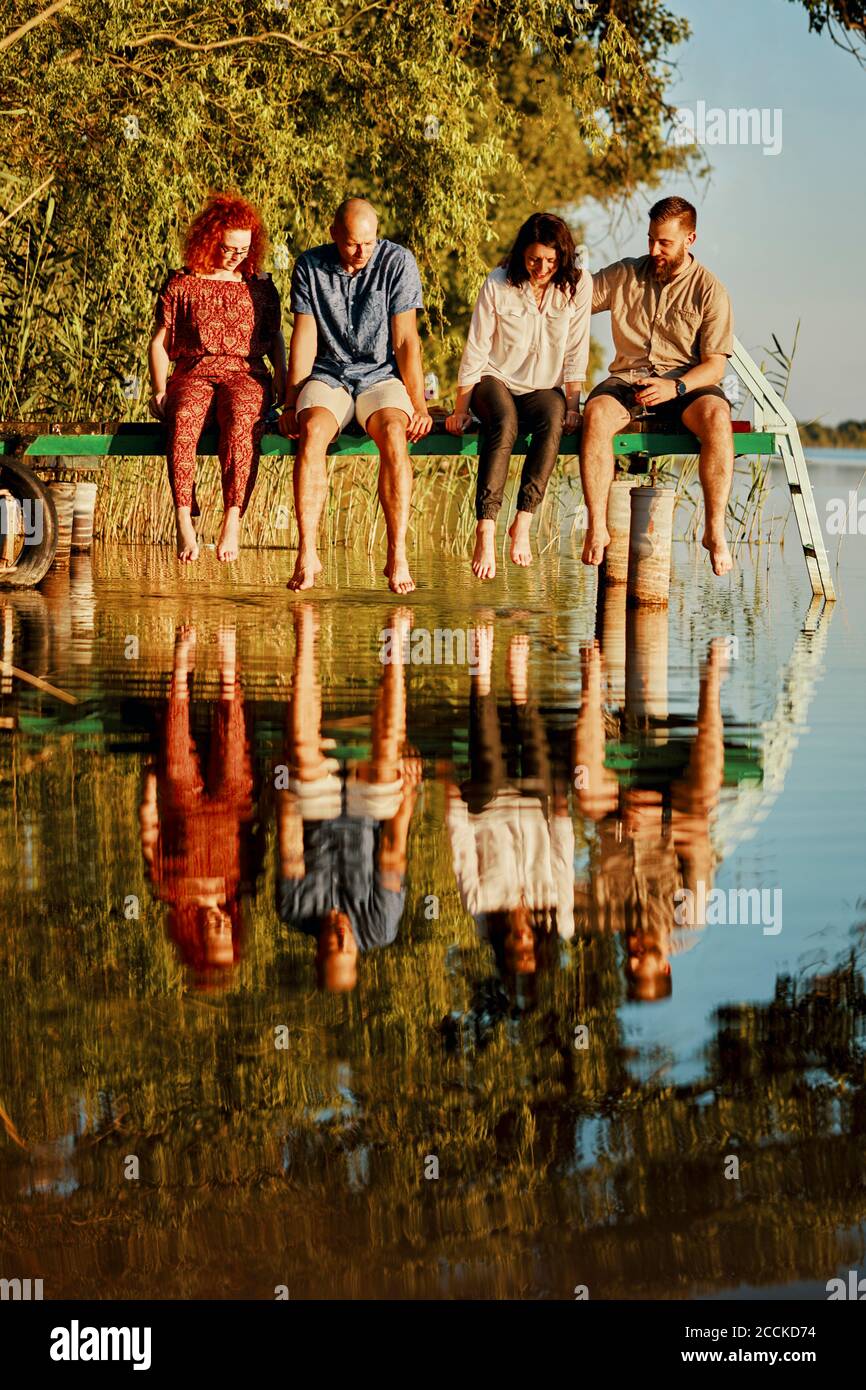 Friends reflected in water sitting on jetty at a lake Stock Photo - Alamy