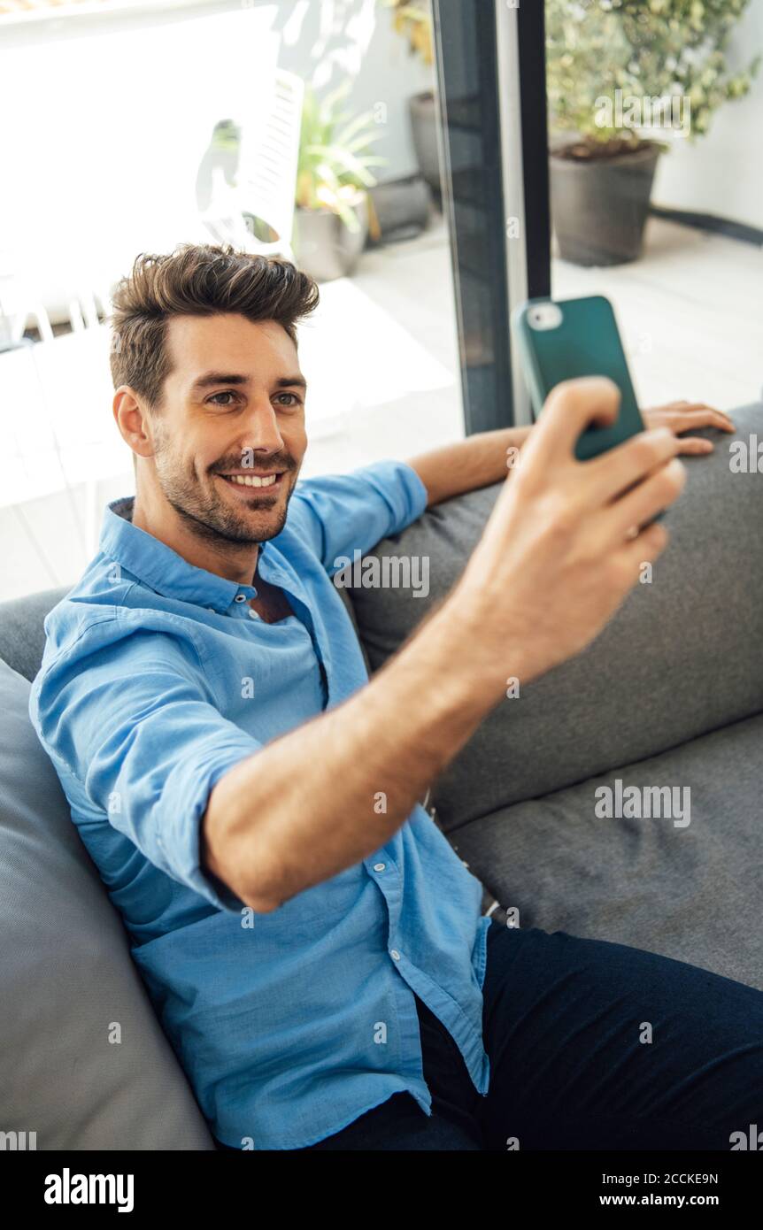 Happy Man Taking Selfie Through Smart Phone While Sitting On Sofa In