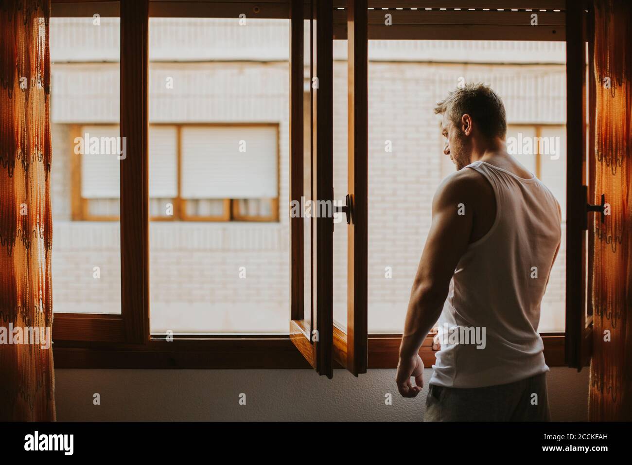 Thoughtful mid adult man looking through window while standing at home ...