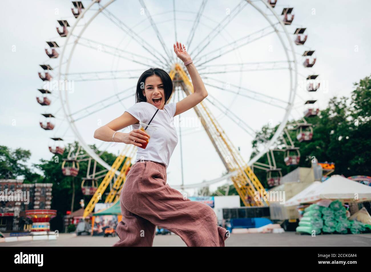 Young woman holding soft drink cup dancing against Ferris wheel in ...