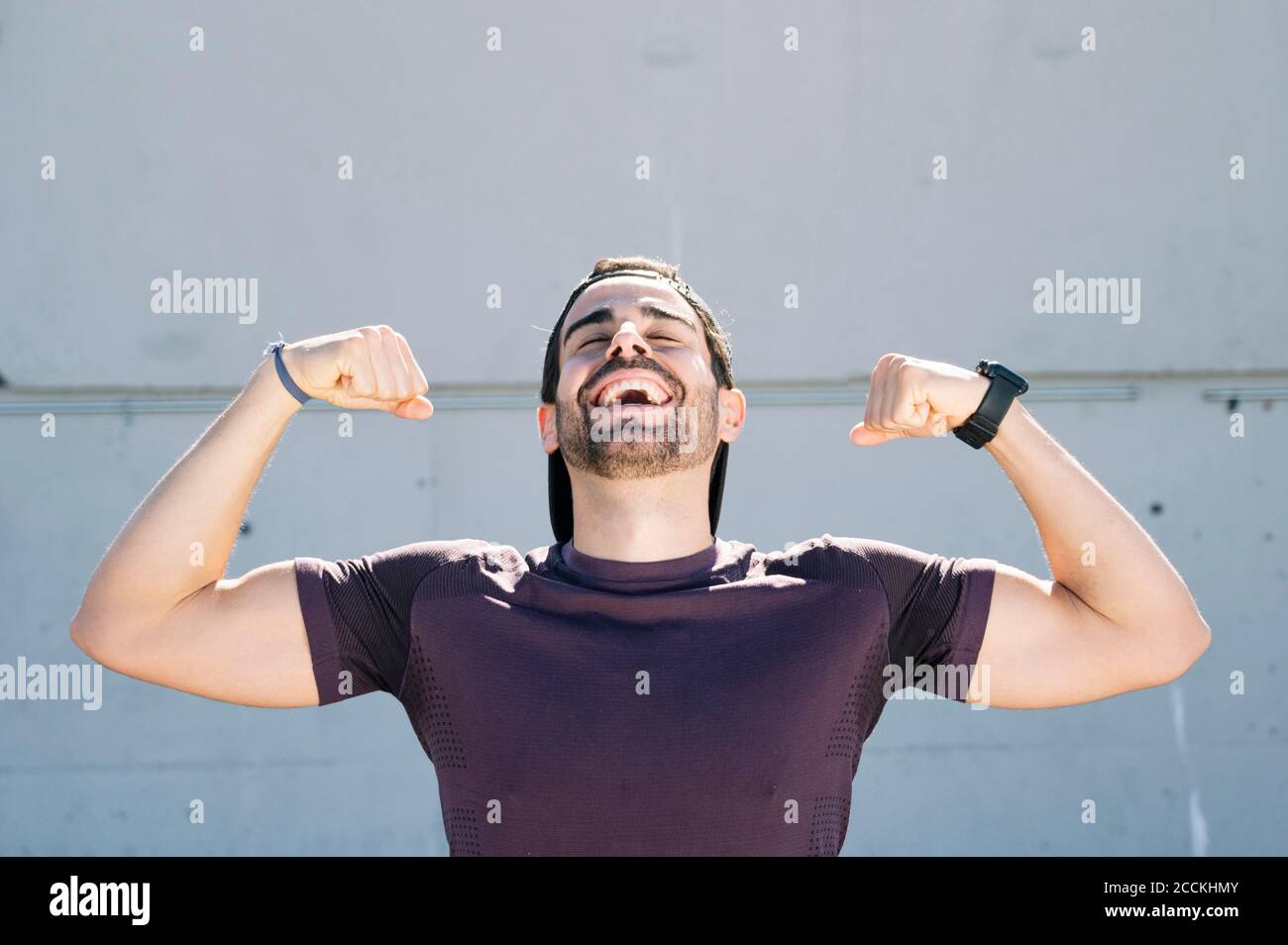 Happy man with eyes closed flexing muscles while standing against wall ...