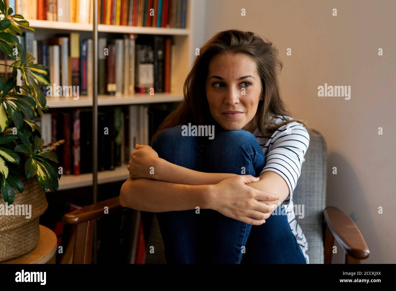 Thoughtful young woman hugging knees while sitting on chair at home ...