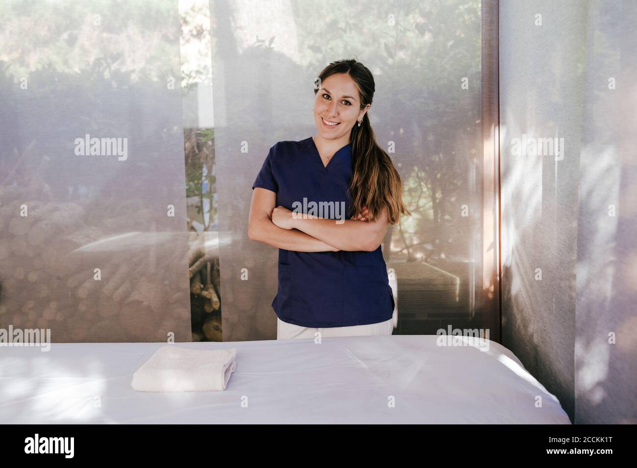 Smiling female massage therapist with arms crossed standing by table in ...