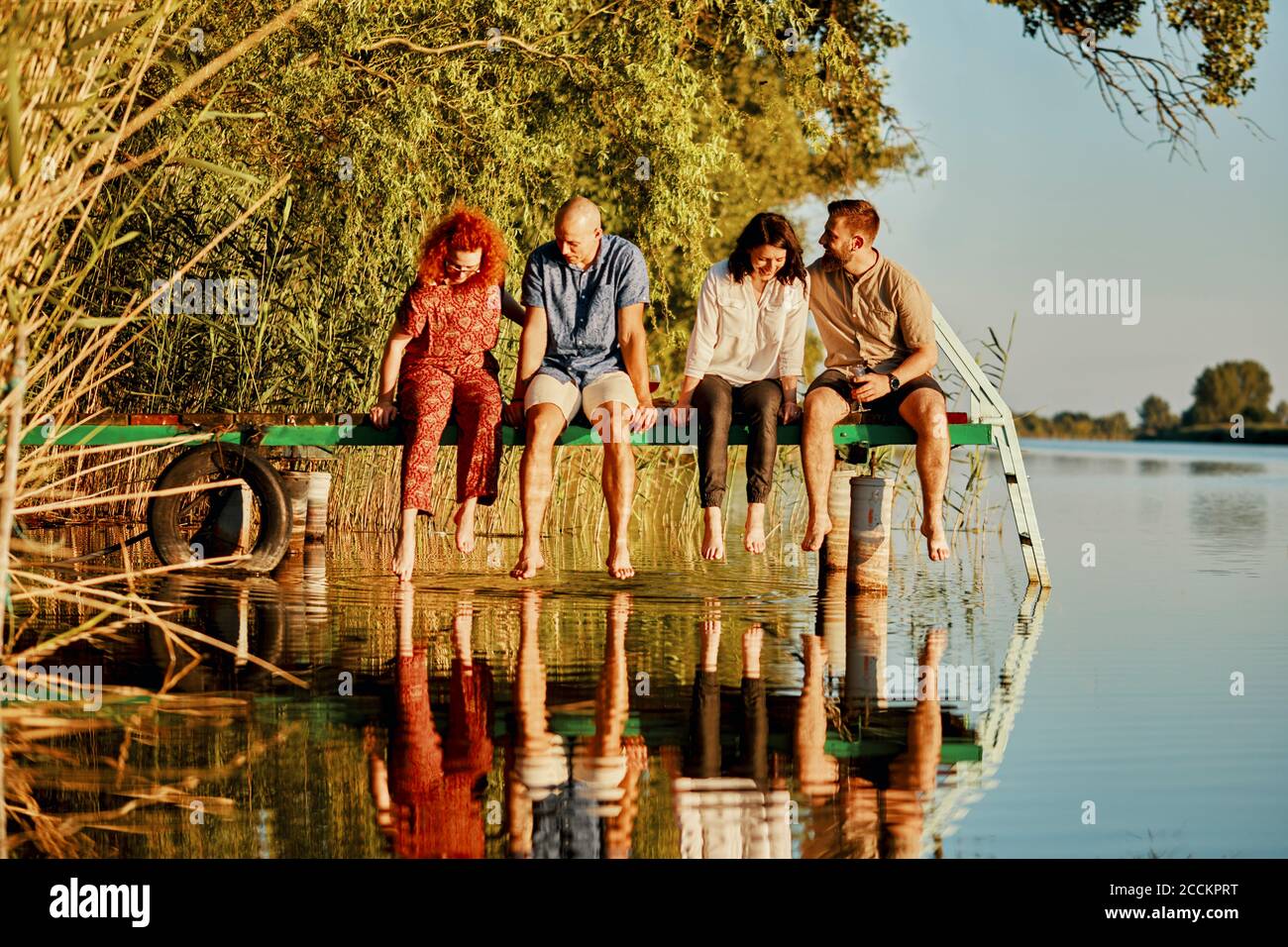 Friends reflected in water sitting on jetty at a lake Stock Photo - Alamy