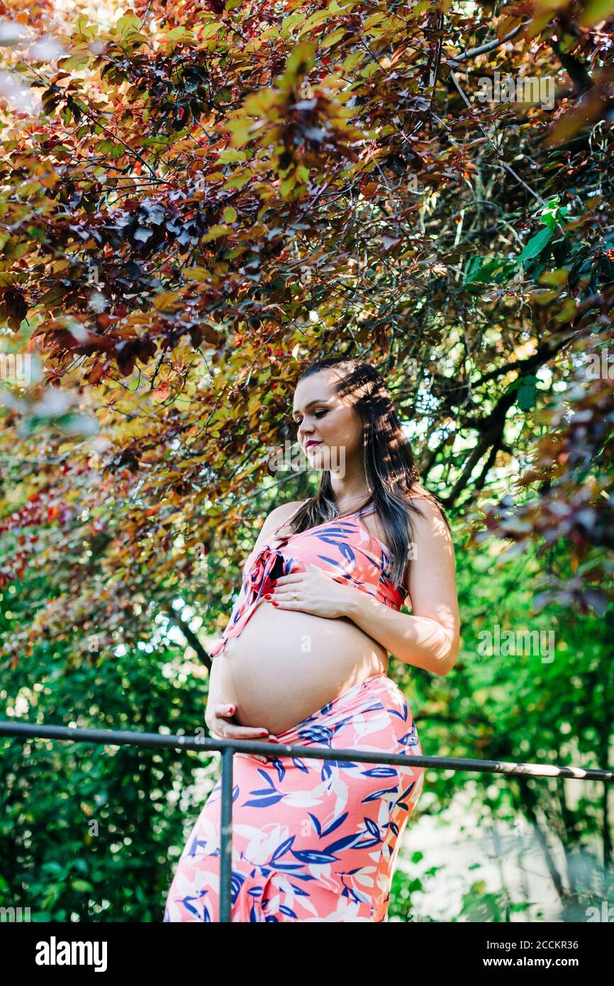 Thoughtful woman standing with hands on stomach below tree in public ...