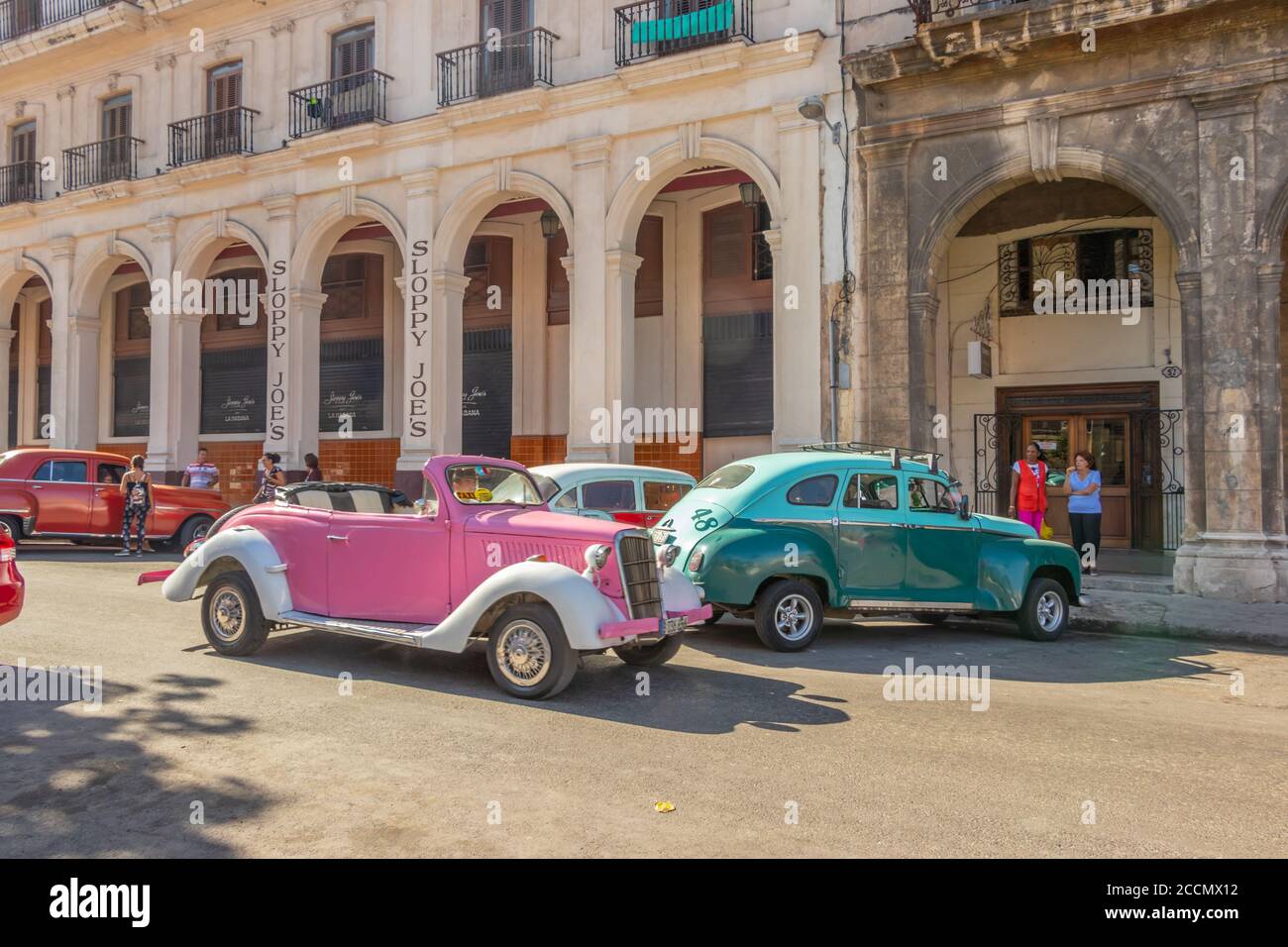 Colorful vintage cars parked on a vibrant street in front of historic buildings with people ...