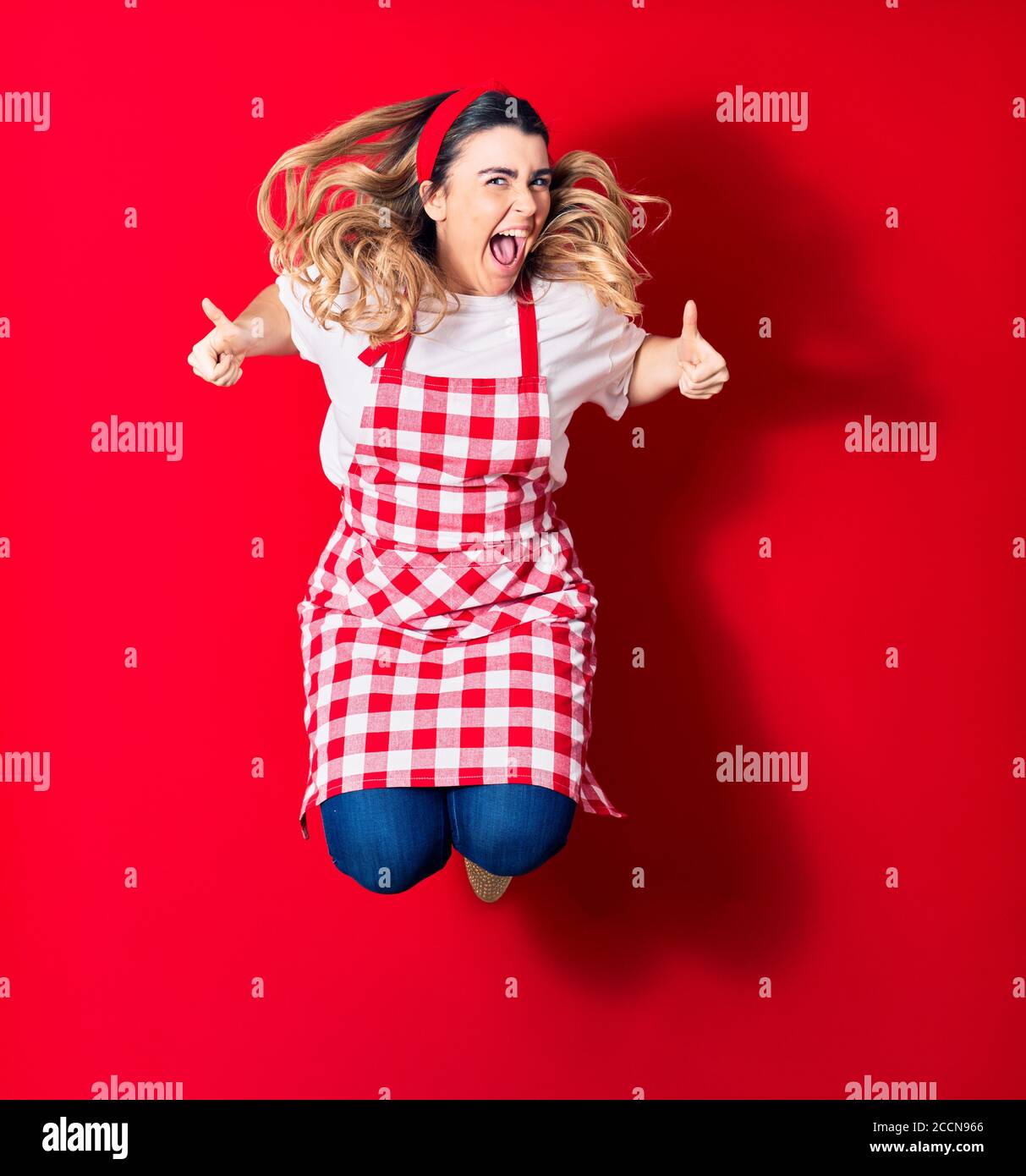 Young beautiful blonde cook woman wearing apron smiling happy. Jumping ...