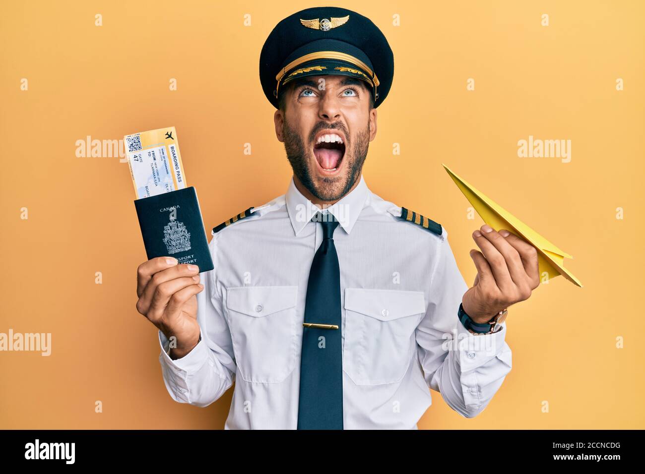 Handsome hispanic pilot man holding paper plane and passport angry and ...