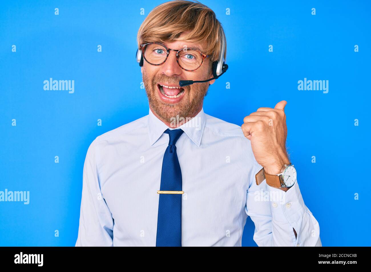 Young blond man wearing call center agent headset pointing thumb up to ...